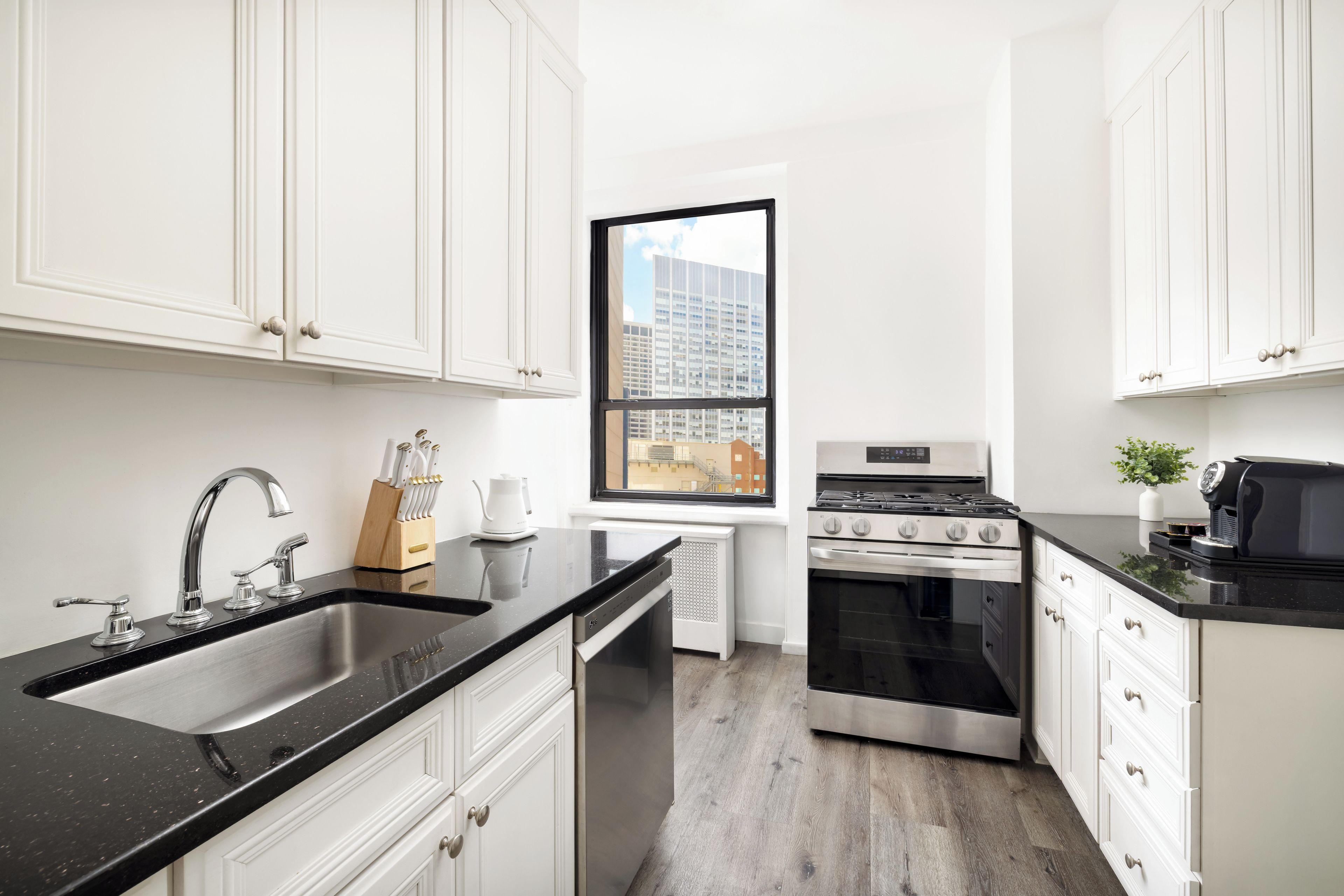 Cozinha moderna com armários brancos, bancada preta, eletrodomésticos de aço inoxidável e janela com vista para a cidade.