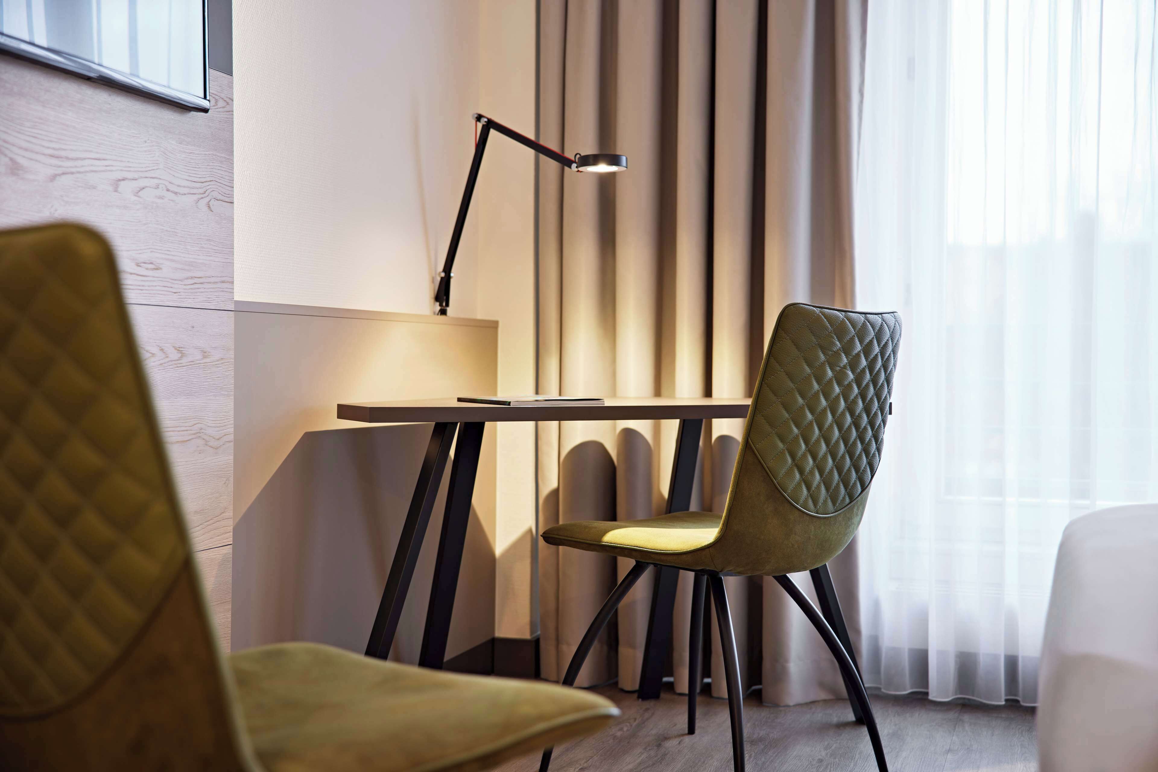 Quarto moderno com piso de madeira, uma escrivaninha com luminária e duas cadeiras verdes.