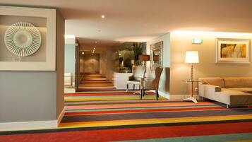 Um hall de hotel com um tapete colorido listrado, um sofá, uma mesa com abajur e um quadro na parede.