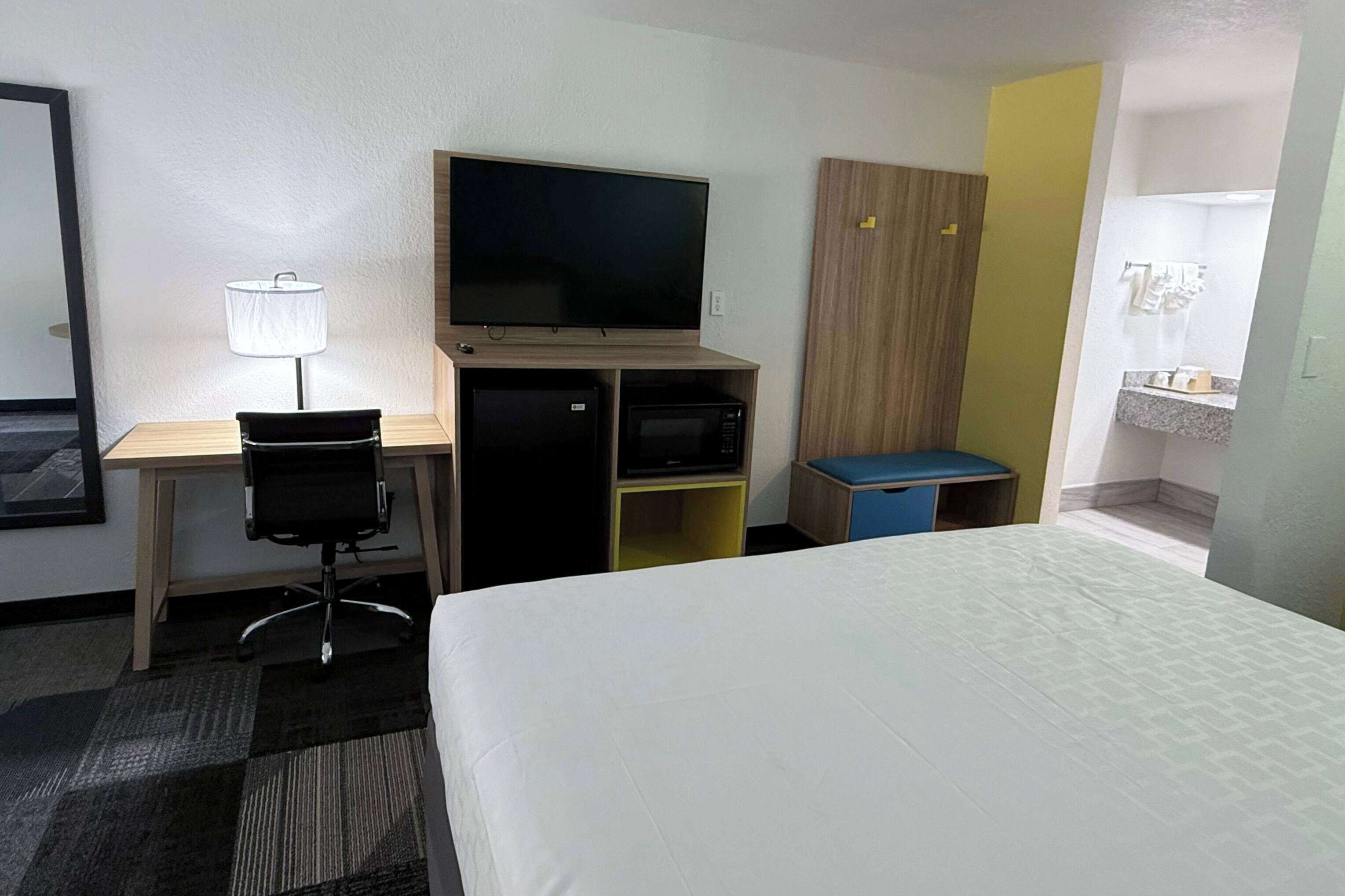 Quarto de hotel com cama, escrivaninha com computador, televisão, armário pequeno e banheiro com espelho e pia.
