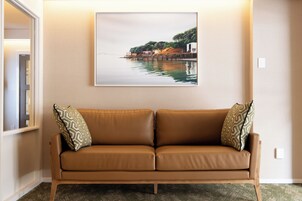 Um sofá de couro bege com almofadas estampadas em frente a uma parede, com um quadro de paisagem emoldurado acima.