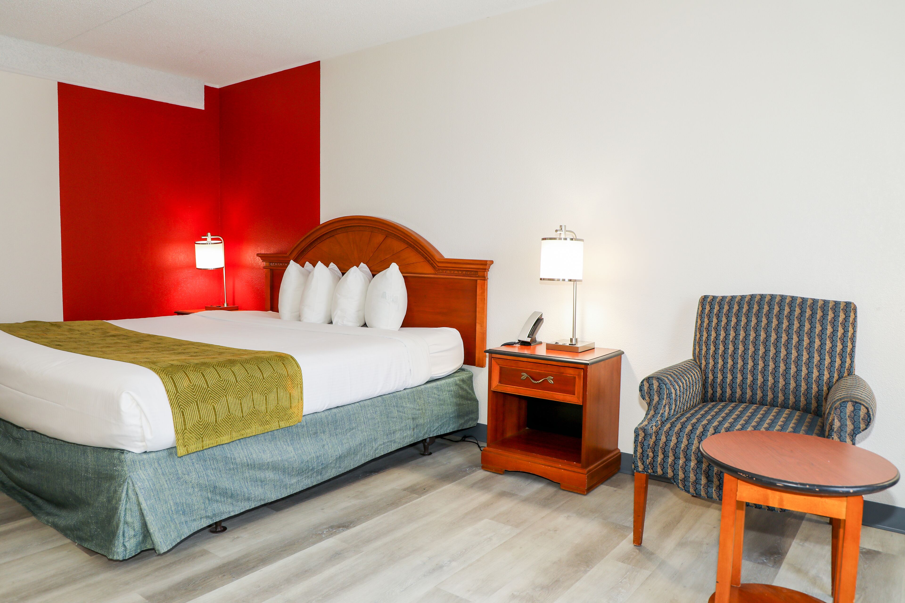 Quarto de hotel com cama, mesa de cabeceira com abajur, cadeira e uma pequena mesa.