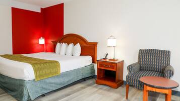 Quarto de hotel com cama, mesa de cabeceira com abajur, cadeira e uma pequena mesa.
