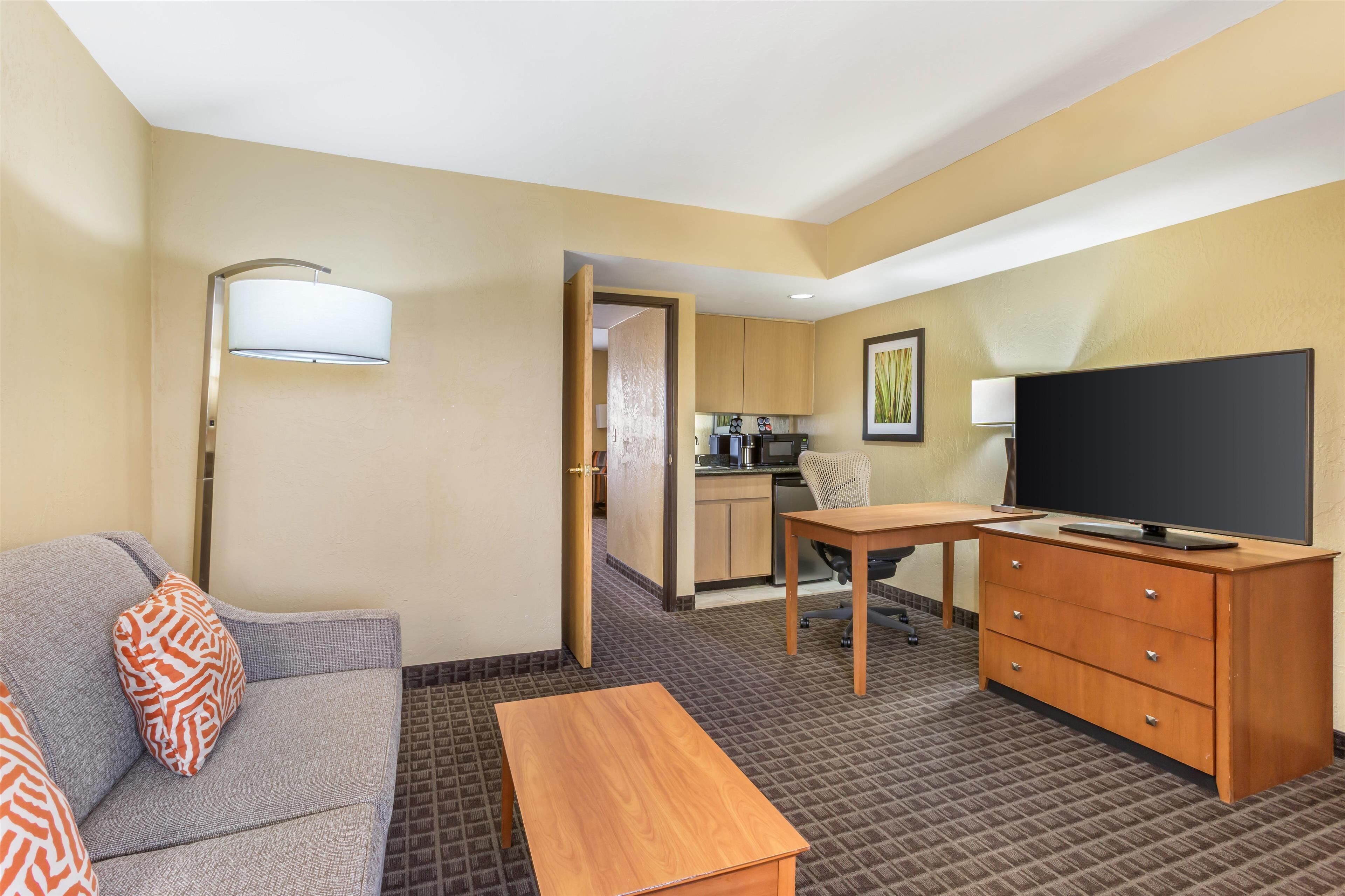 Quarto de hotel com sofá, escrivaninha de madeira, televisão e cozinha americana ao fundo.