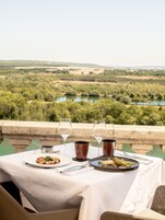 Mesa posta com pratos de comida, copos e um jarro de metal, com vista para uma paisagem cênica com rio e vegetação.