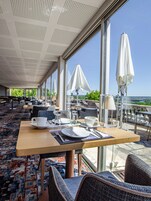 Um restaurante com terraço, mesas preparadas com pratos, talheres e copos, e amplas janelas com vista.