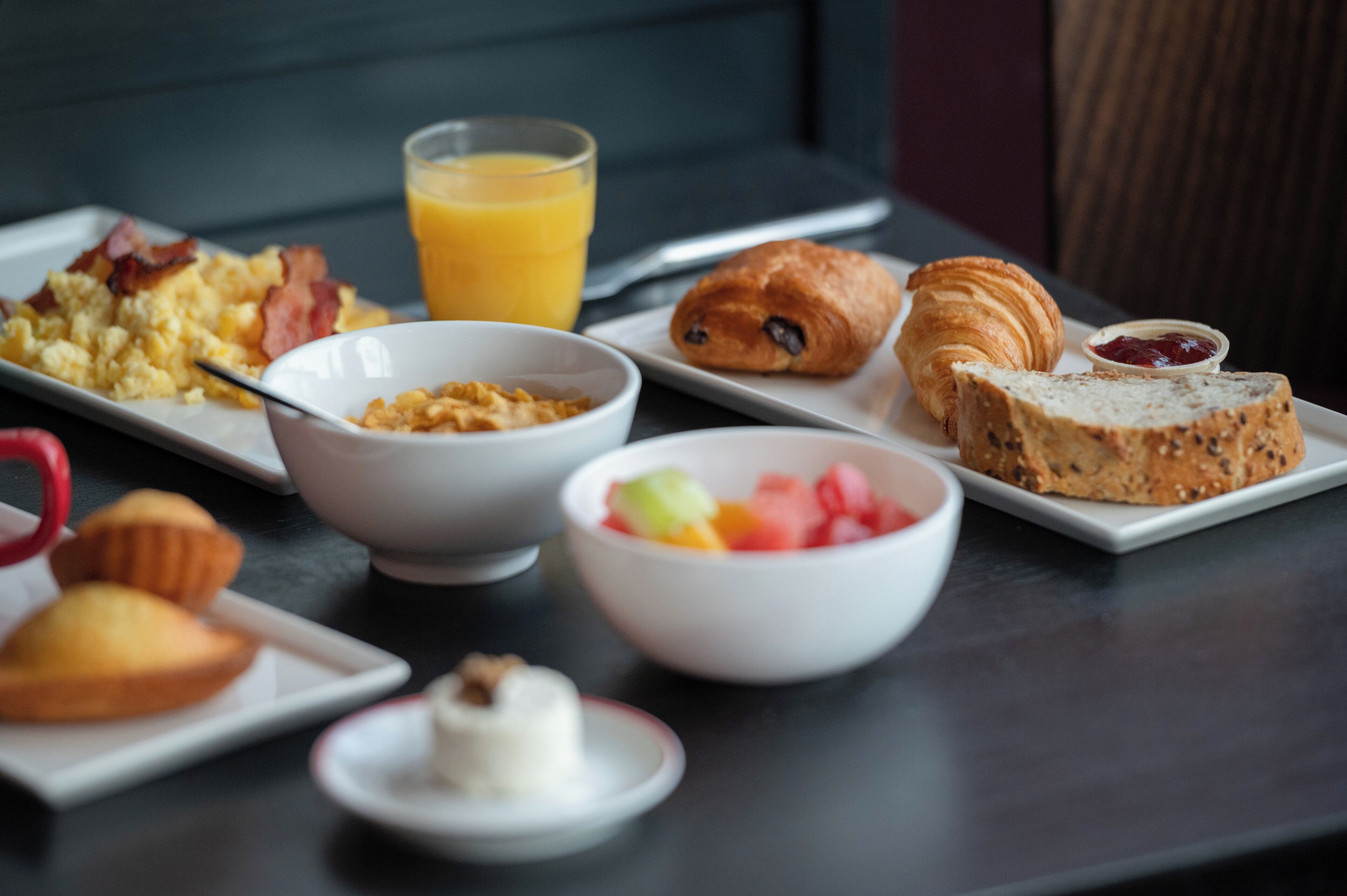 Uma mesa de café da manhã com diversos pratos, incluindo ovos mexidos, bacon, croissants e frutas.