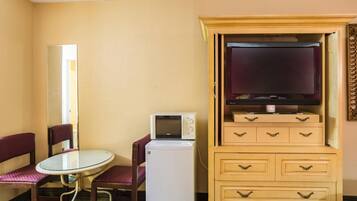 Quarto de hotel compacto com uma mesa redonda, duas cadeiras, um frigorífico branco pequeno, uma televisão num móvel de madeira e um espelho na parede.