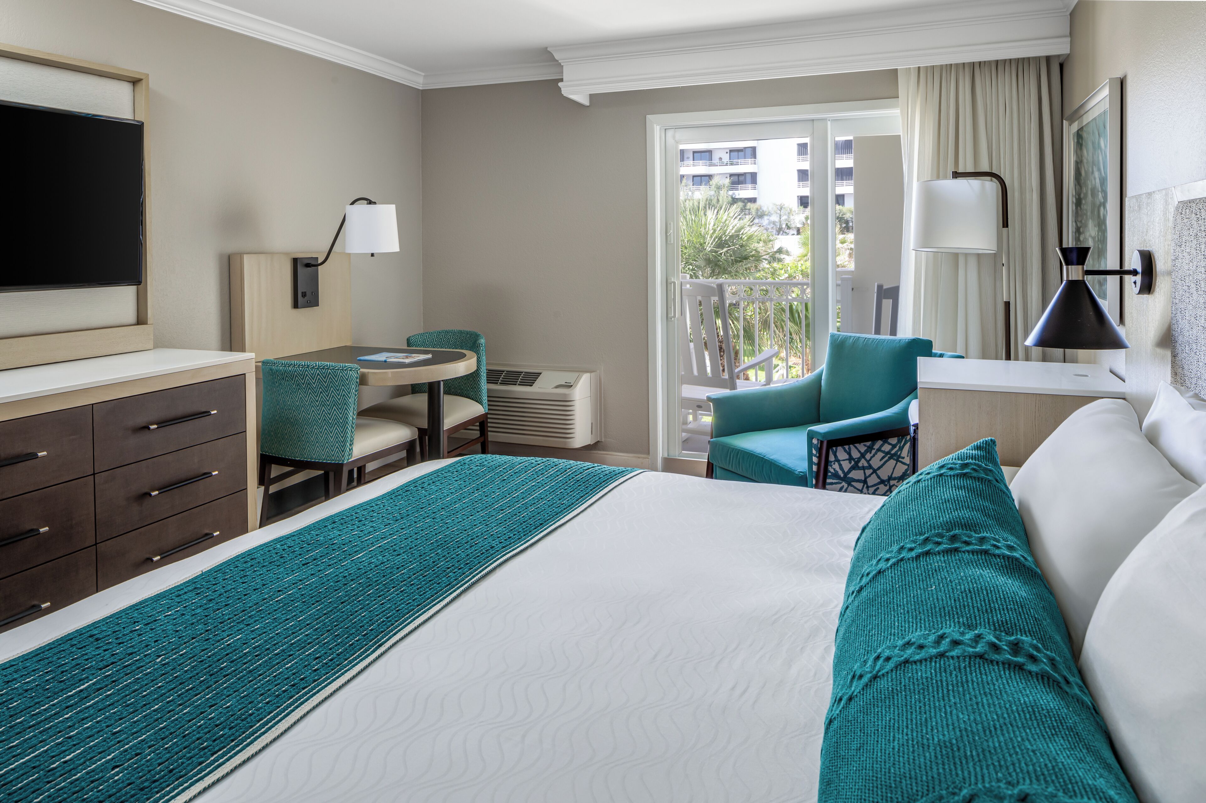 Quarto de hotel com cama, secretária, cadeira, televisão e varanda com vista para edifícios e árvores.