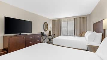 Quarto de hotel com uma cama grande, uma cama menor, uma escrivaninha com cadeira, televisão e uma janela com cortinas.