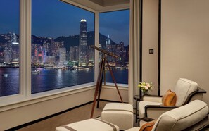 Quarto de hotel com uma grande janela com vista para a paisagem urbana à noite, com um telescópio e área de estar.