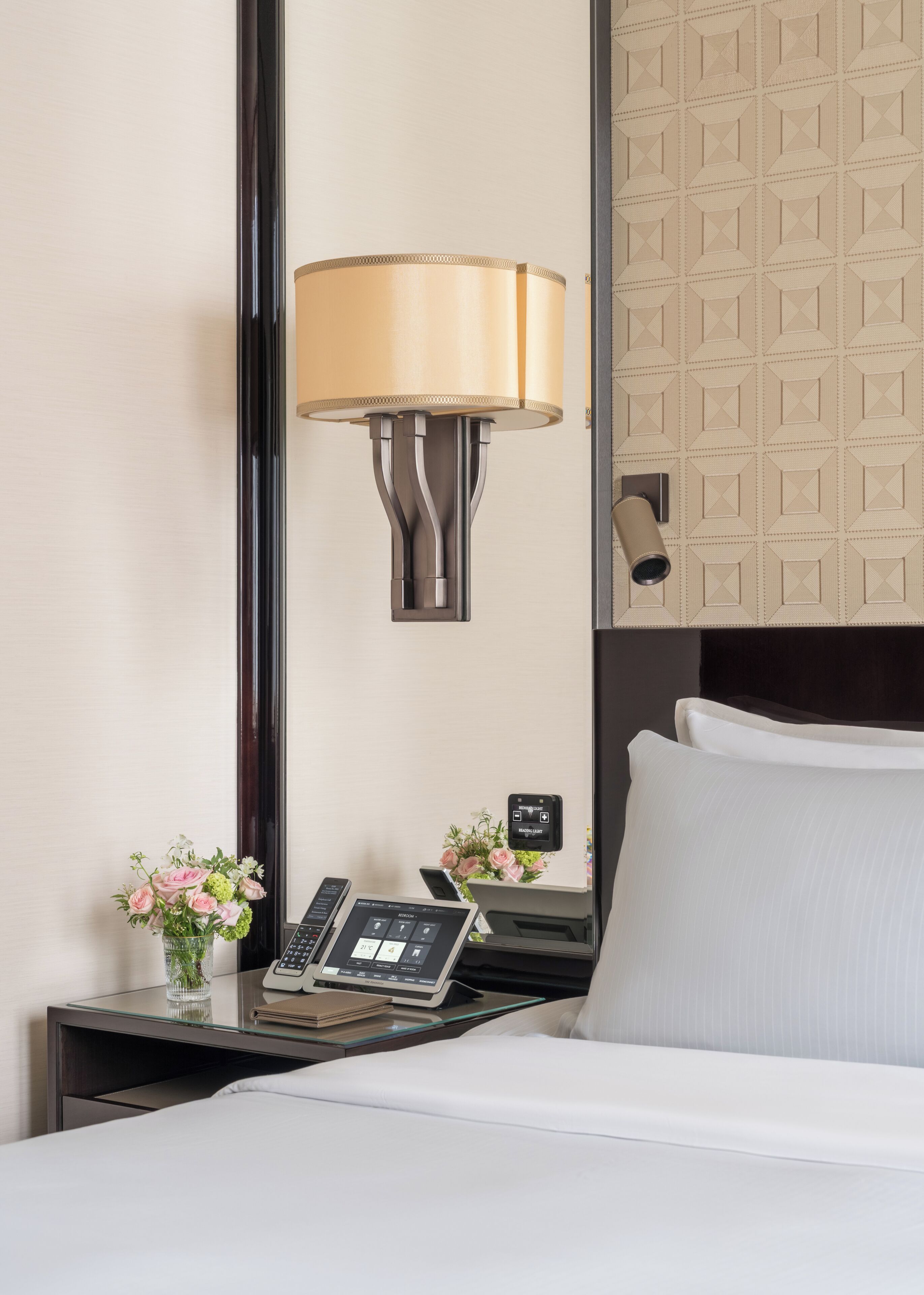 Quarto de hotel com cama, mesa de cabeceira com luminária, telefone, pequeno vaso com flores e painel de controle para os serviços do quarto.