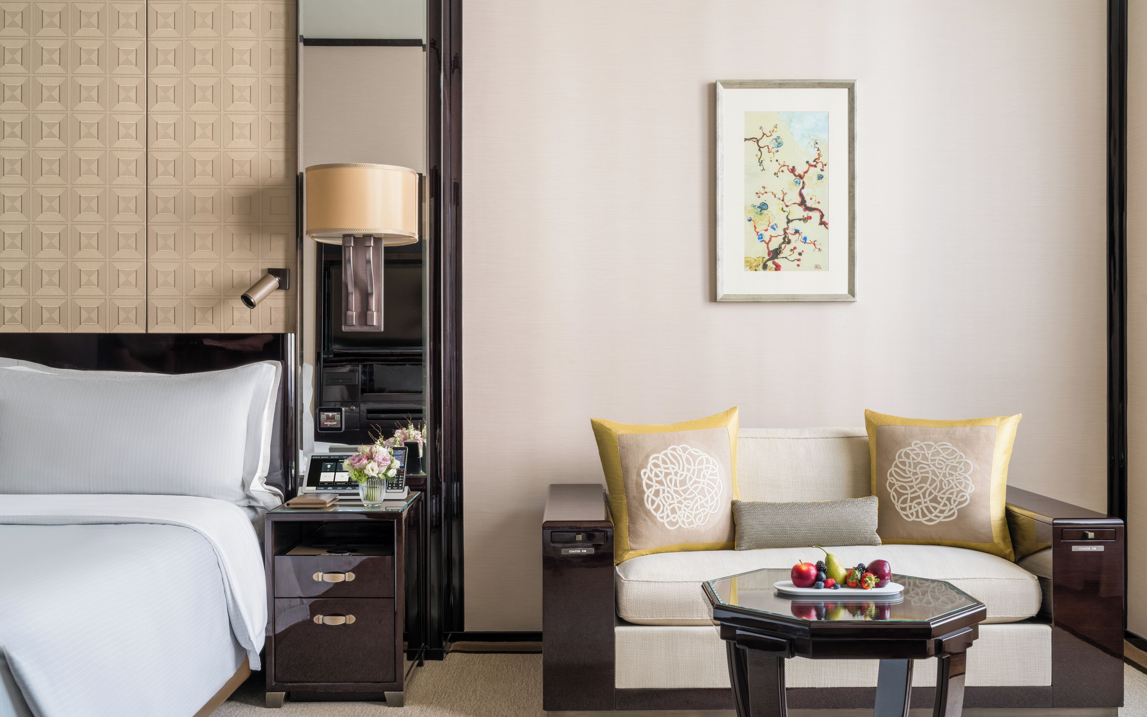 Quarto de hotel com cama, mesa de cabeceira com abajur, sofá com almofadas decorativas, pequena mesa com frutas e uma obra de arte emoldurada na parede.