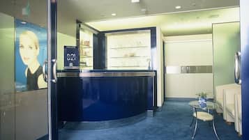 Interior moderno com balcão de recepção, porta de vidro com um cartaz, pequena mesa com uma planta e área de estar com sofá e poltronas.