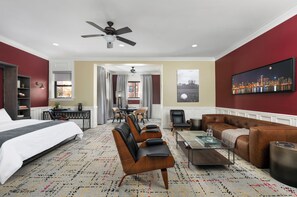 Quarto de hotel com cama, área de estar com poltronas de couro, mesa de centro, televisão e pintura com vista urbana na parede.