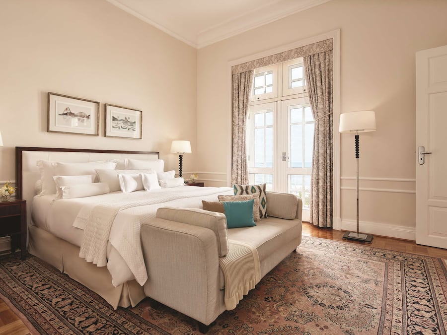 Quarto com uma cama grande, um sofá, uma mesa de cabeceira, um abajur, uma janela com cortinas e uma porta.