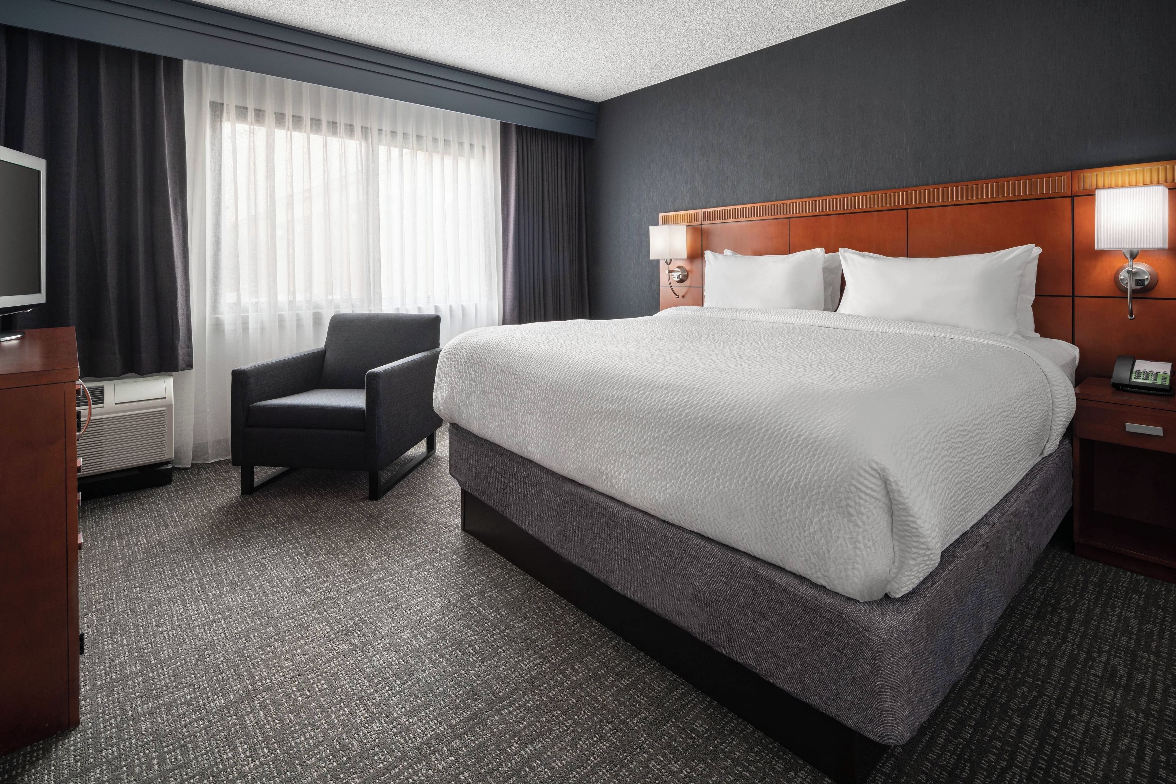 Quarto de hotel com uma cama grande, uma cadeira, escrivaninha com telefone e janela com cortinas.