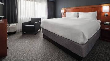 Quarto de hotel com uma cama grande, uma cadeira, escrivaninha com telefone e janela com cortinas.