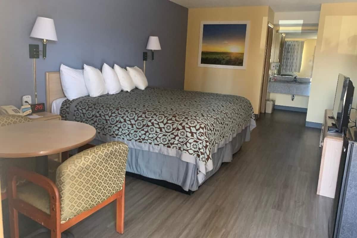 Quarto de hotel com cama, abajures, escrivaninha com cadeira, televisão e um quadro na parede.