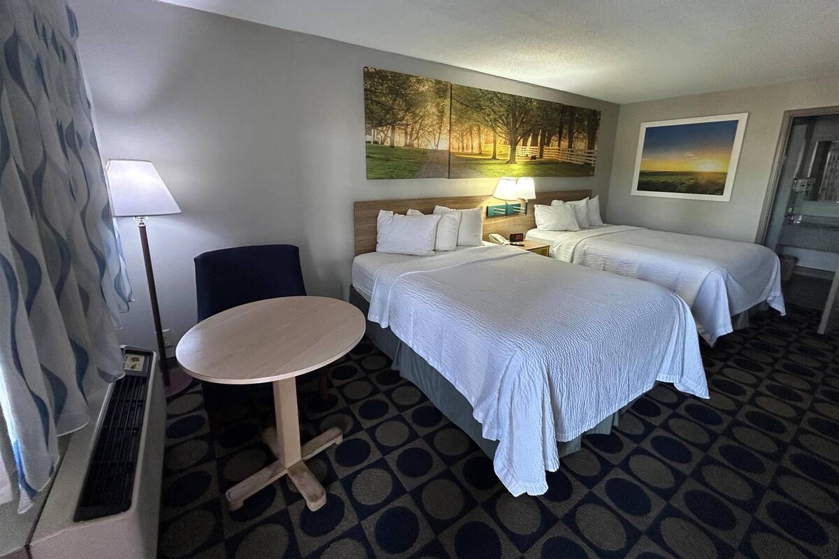 Quarto de hotel com duas camas, uma mesinha, um abajur e um quadro na parede.