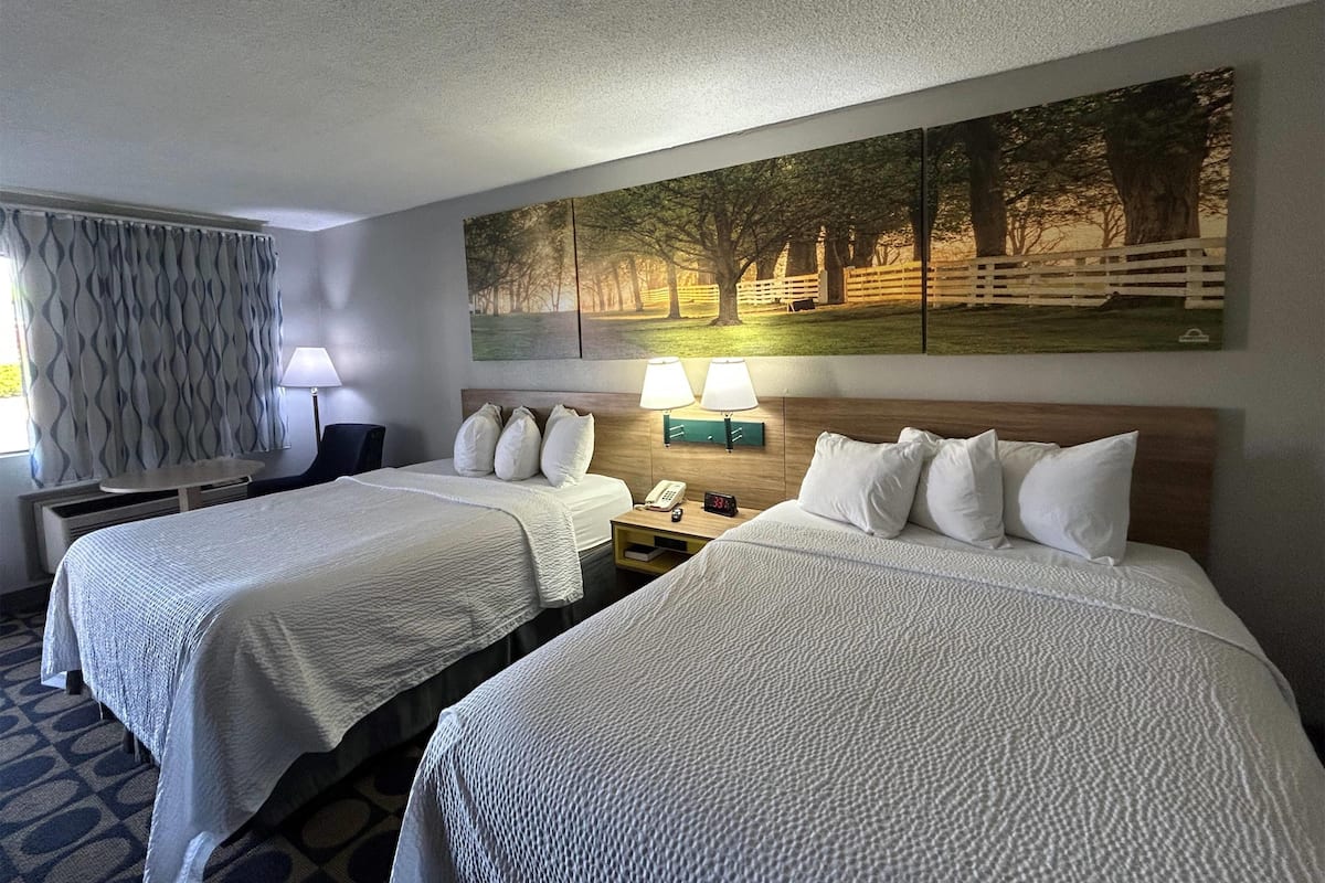 Quarto de hotel com duas camas, mesa de cabeceira, abajur e pintura de árvores.