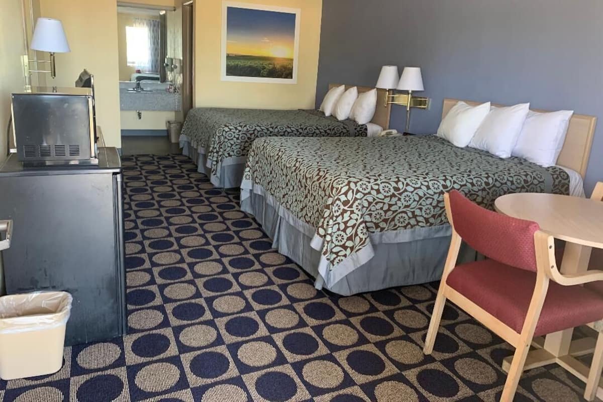 Quarto de hotel com duas camas, uma escrivaninha, uma cadeira, uma TV e um quadro na parede.