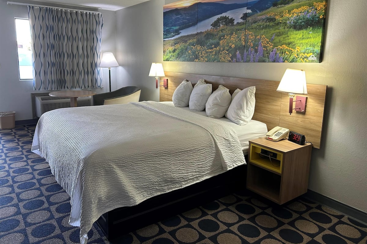 Quarto de hotel com uma cama grande, duas luminárias de cabeceira, mesa de cabeceira com telefone e relógio, escrivaninha com cadeira e janela com cortinas.