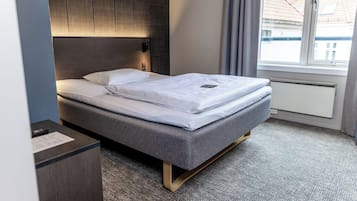 Uma cama bem arrumada com colchão cinza e lençóis brancos, uma mesa de cabeceira com controle remoto e uma janela com vista para edifícios.