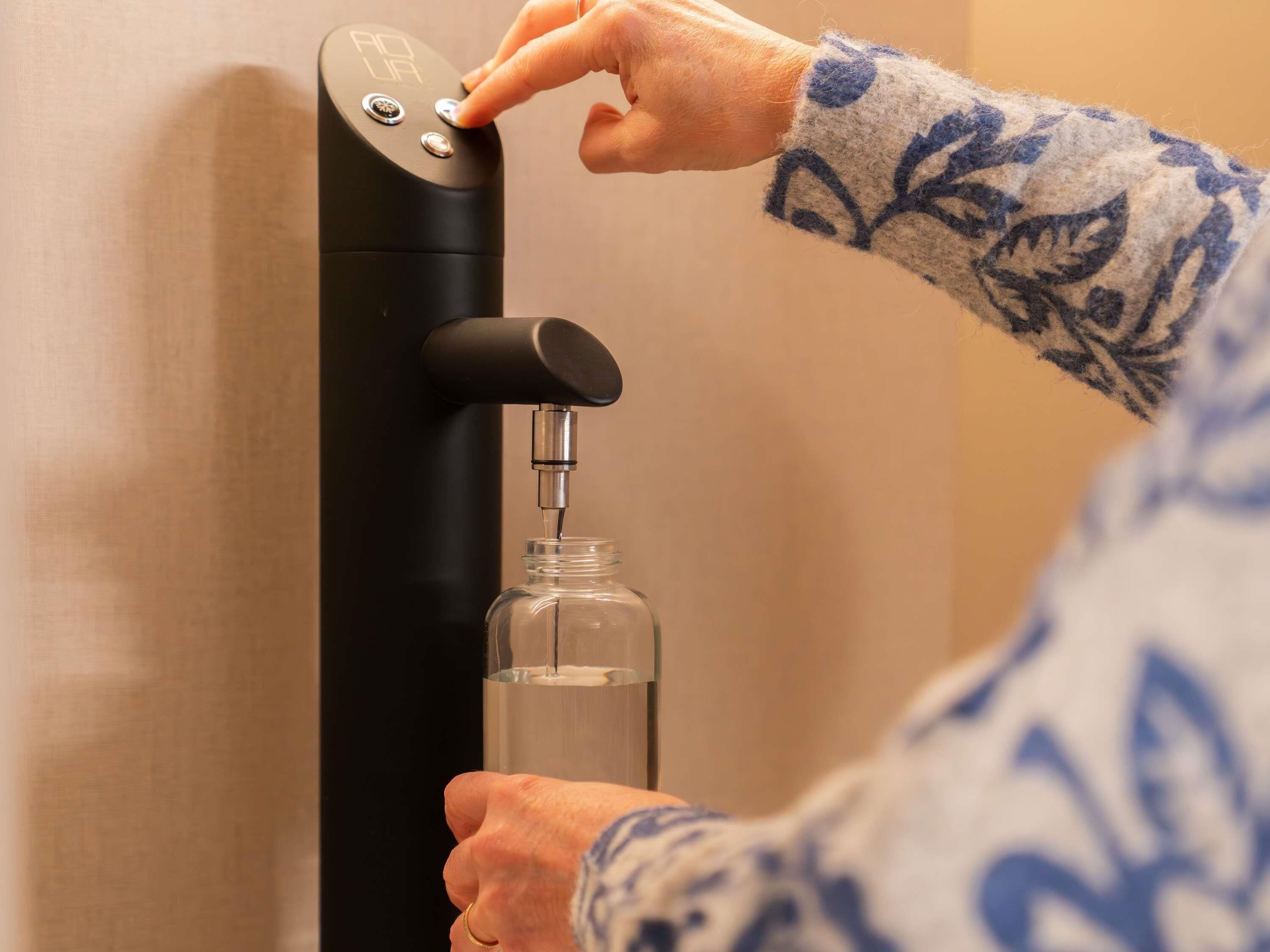 Uma pessoa utilizando um dispensador preto fixado na parede, com um frasco transparente, provavelmente para sabonete ou loção.