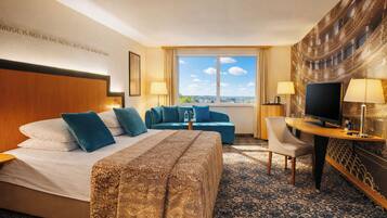 Quarto de hotel com cama, dois sofás, escrivaninha com televisão e grande janela com vista para a cidade.