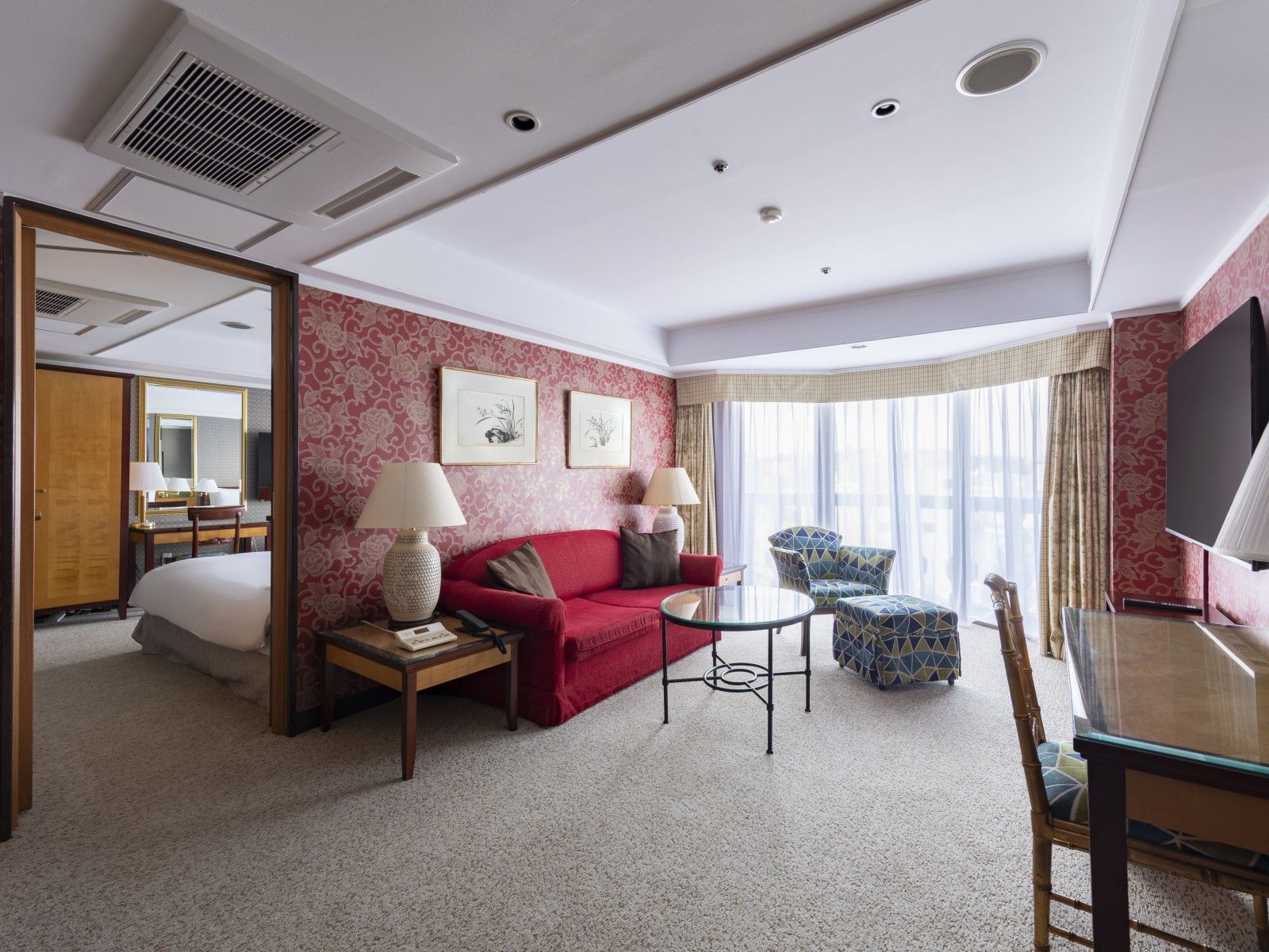 Quarto de hotel com cama, sofá vermelho, mesa de centro, televisão e uma janela grande com cortinas.