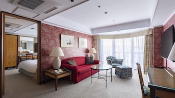 Quarto de hotel com cama, sofá vermelho, mesa de centro, televisão e uma janela grande com cortinas.