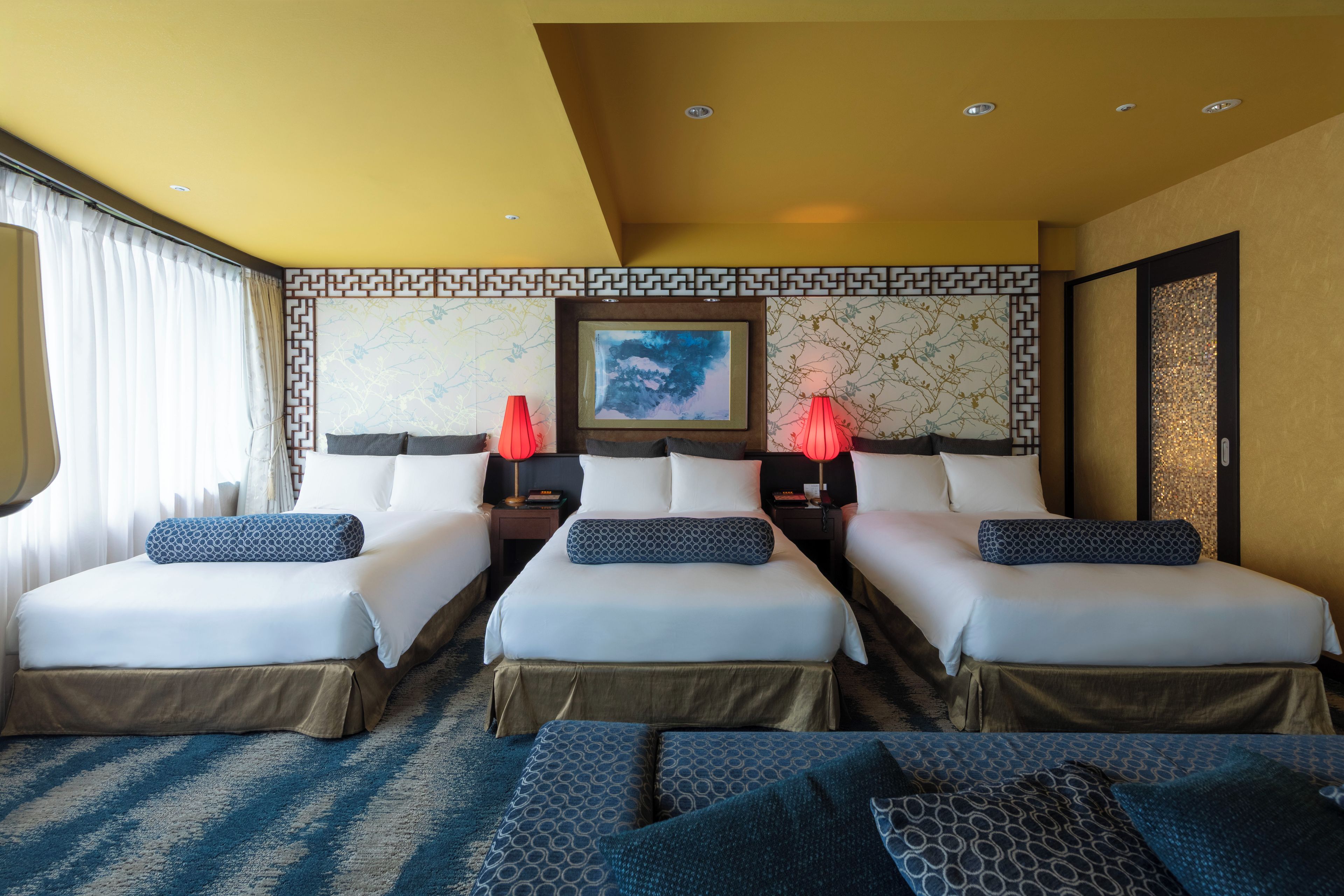 Quarto de hotel com três camas, uma janela grande com cortinas, um painel decorativo na parede e um abajur vermelho.