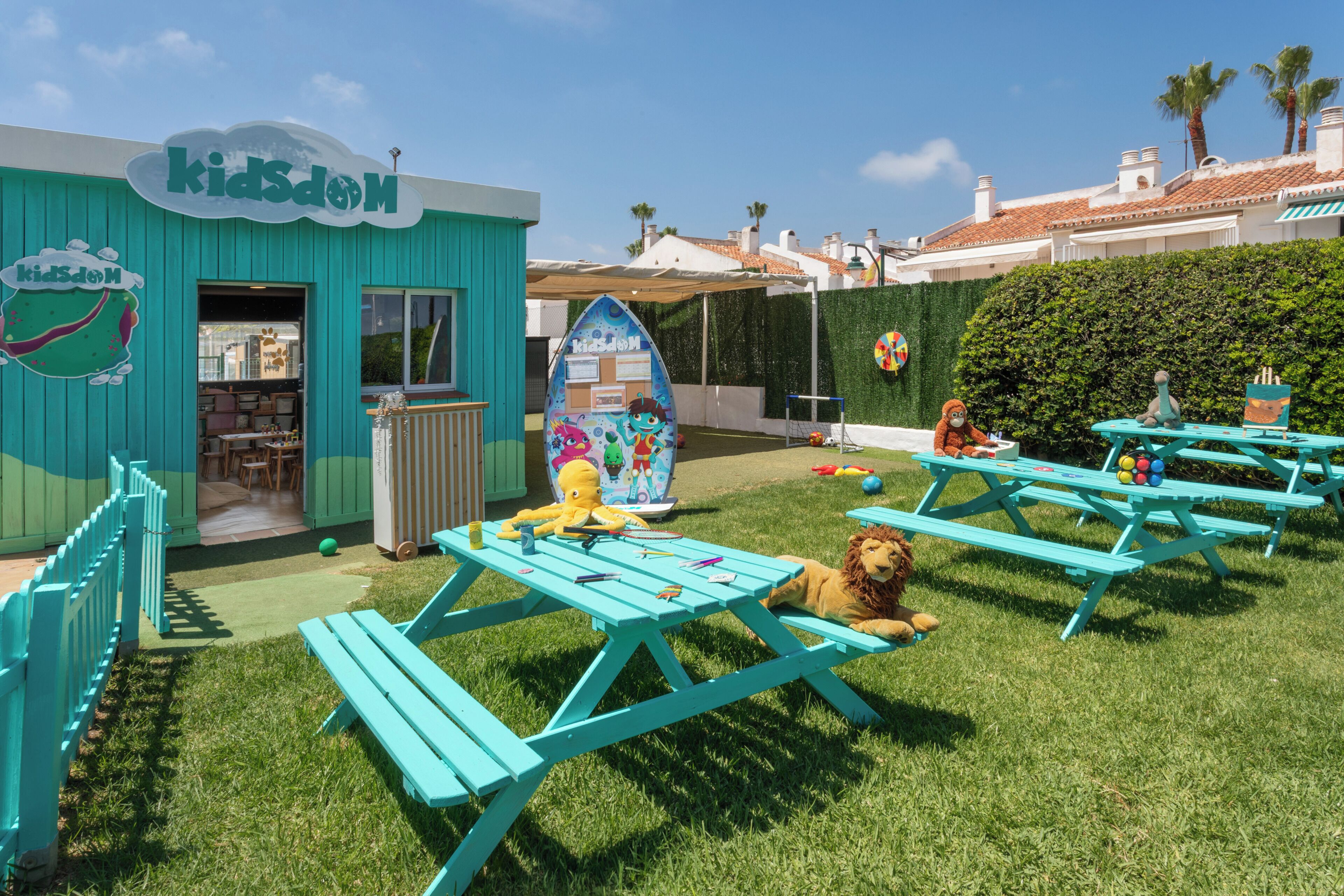 Uma área de recreação infantil com um prédio azul identificado como 'kidsdom', com mesas de piquenique, um pequeno gol de futebol e diversos brinquedos.