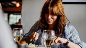 Uma pessoa jantando à mesa com um prato de comida e um copo de vinho branco.