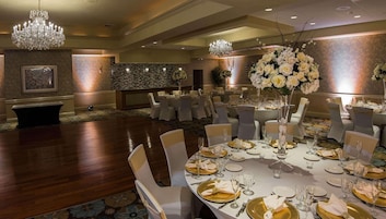 Uma sala de banquetes com mesas redondas dispostas para um evento formal, com lustres, centros de mesa florais e parede estampada.