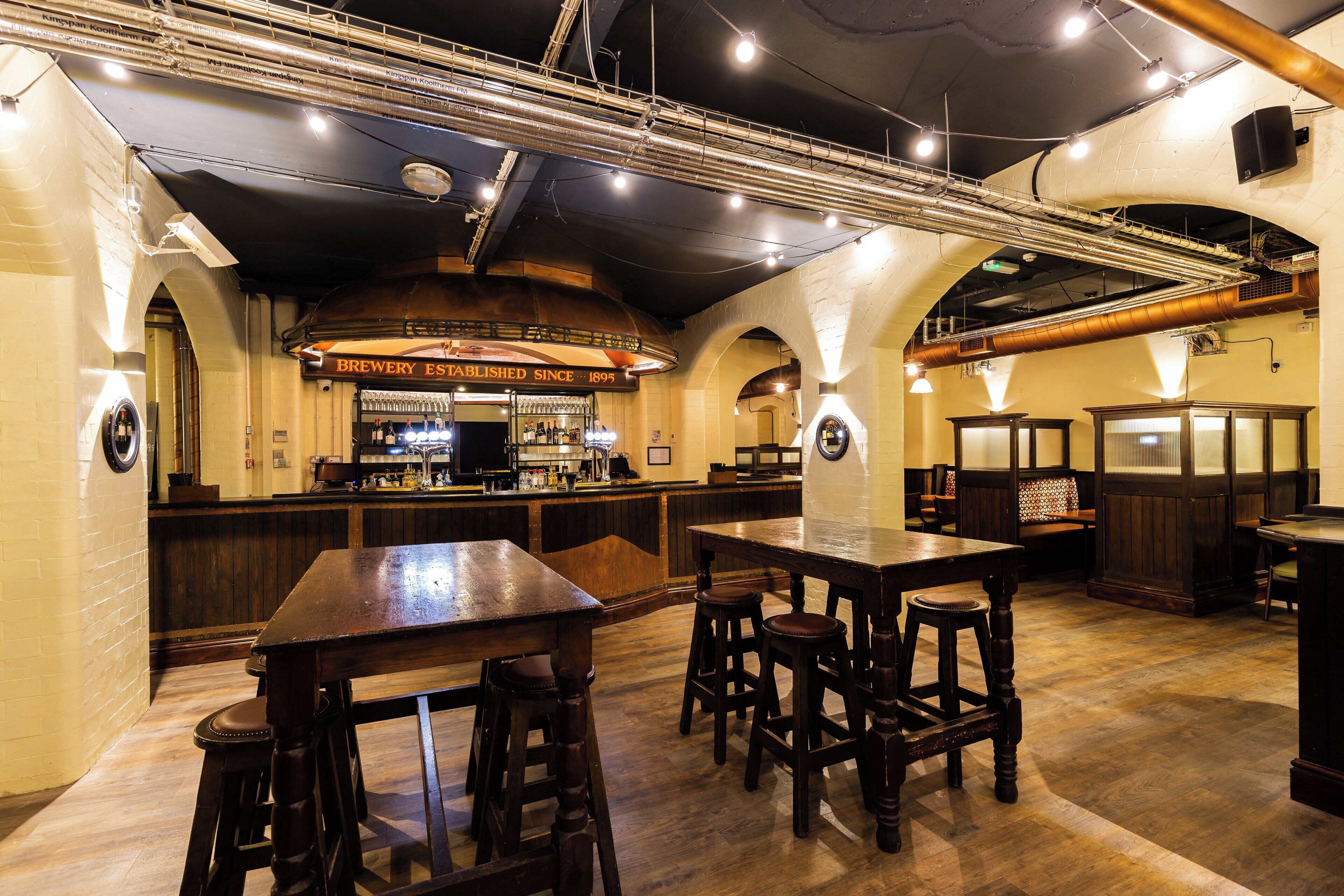 Uma área de bar com mesas e banquetas de madeira, um balcão com a placa 'BREWERY ESTABLISHED SINCE 1895' e dutos expostos no teto.