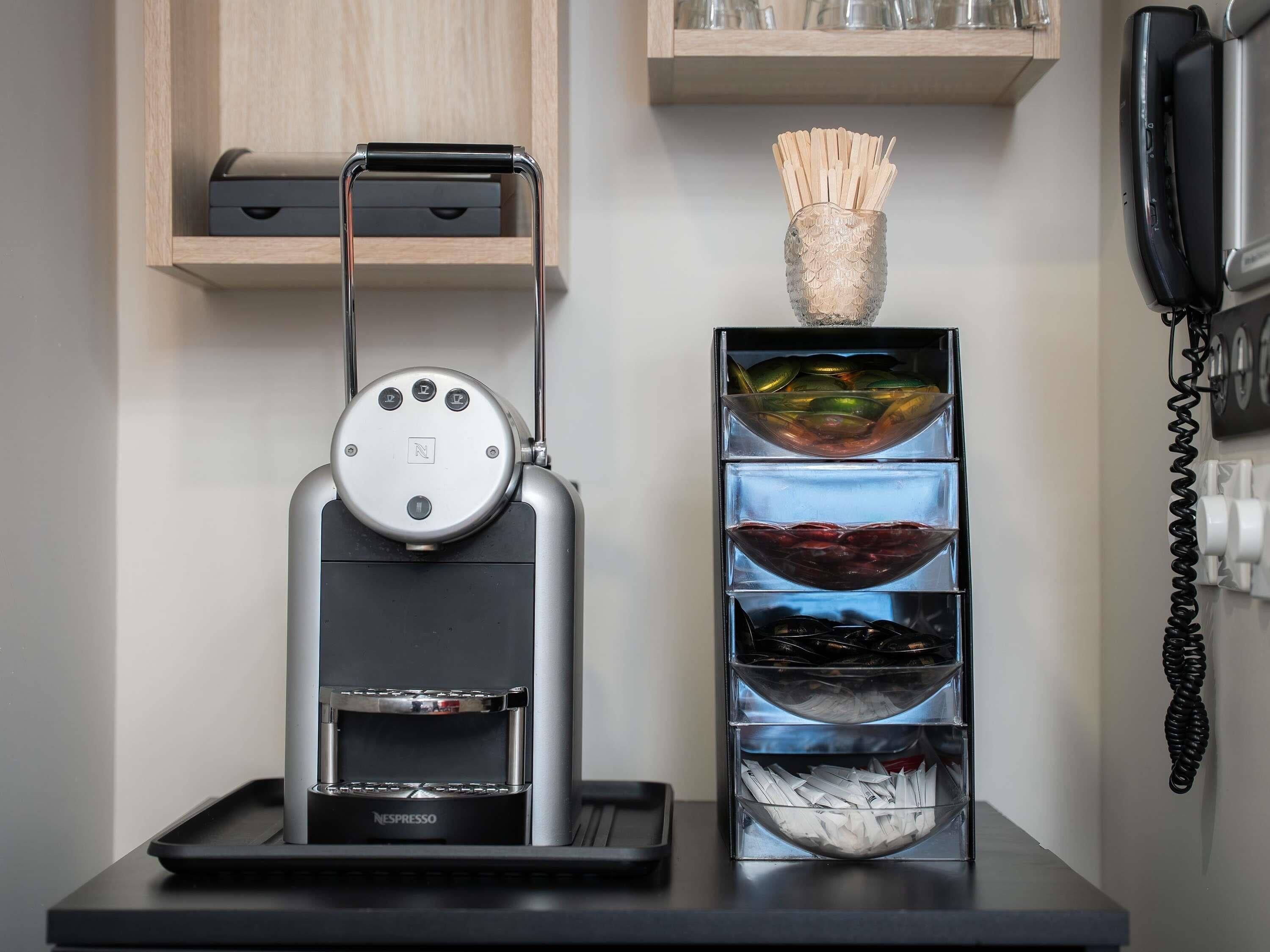 Uma máquina de café Nespresso com reservatório de água, um recipiente de vidro com cabo de madeira e um celular sobre a mesa.