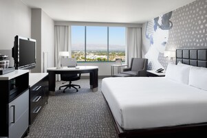 Quarto de hotel com uma cama grande, uma escrivaninha com computador, televisão e vista para a cidade através de amplas janelas.