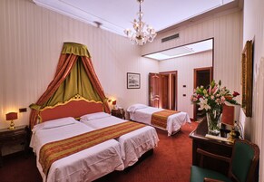 Quarto de hotel com duas camas, um candelabro, uma escrivaninha com uma cadeira e um espelho.