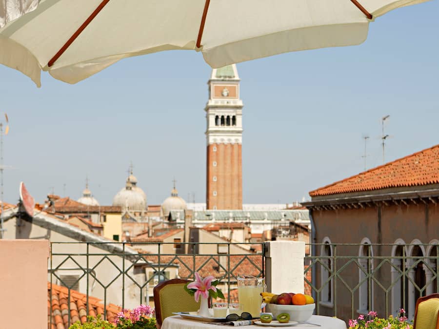 Terraço com mesa posta para refeição, um guarda-sol branco e vista para uma torre histórica.