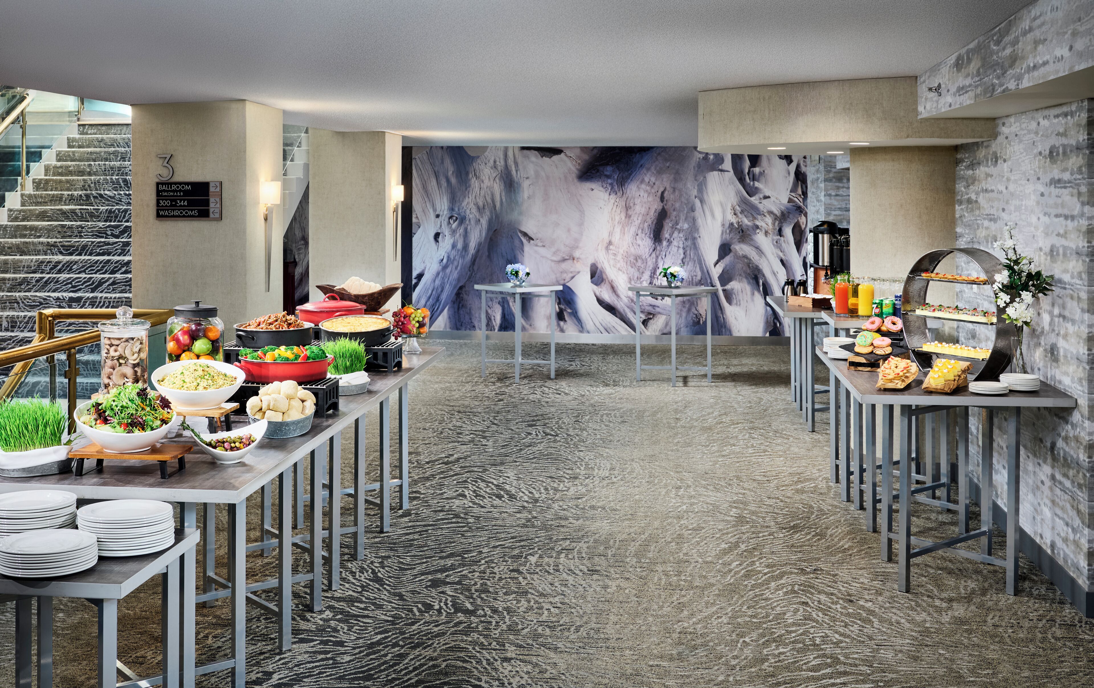 Um quarto amplo com escadaria de mármore, um grande mural na parede e uma mesa de buffet com diversos alimentos e bebidas.