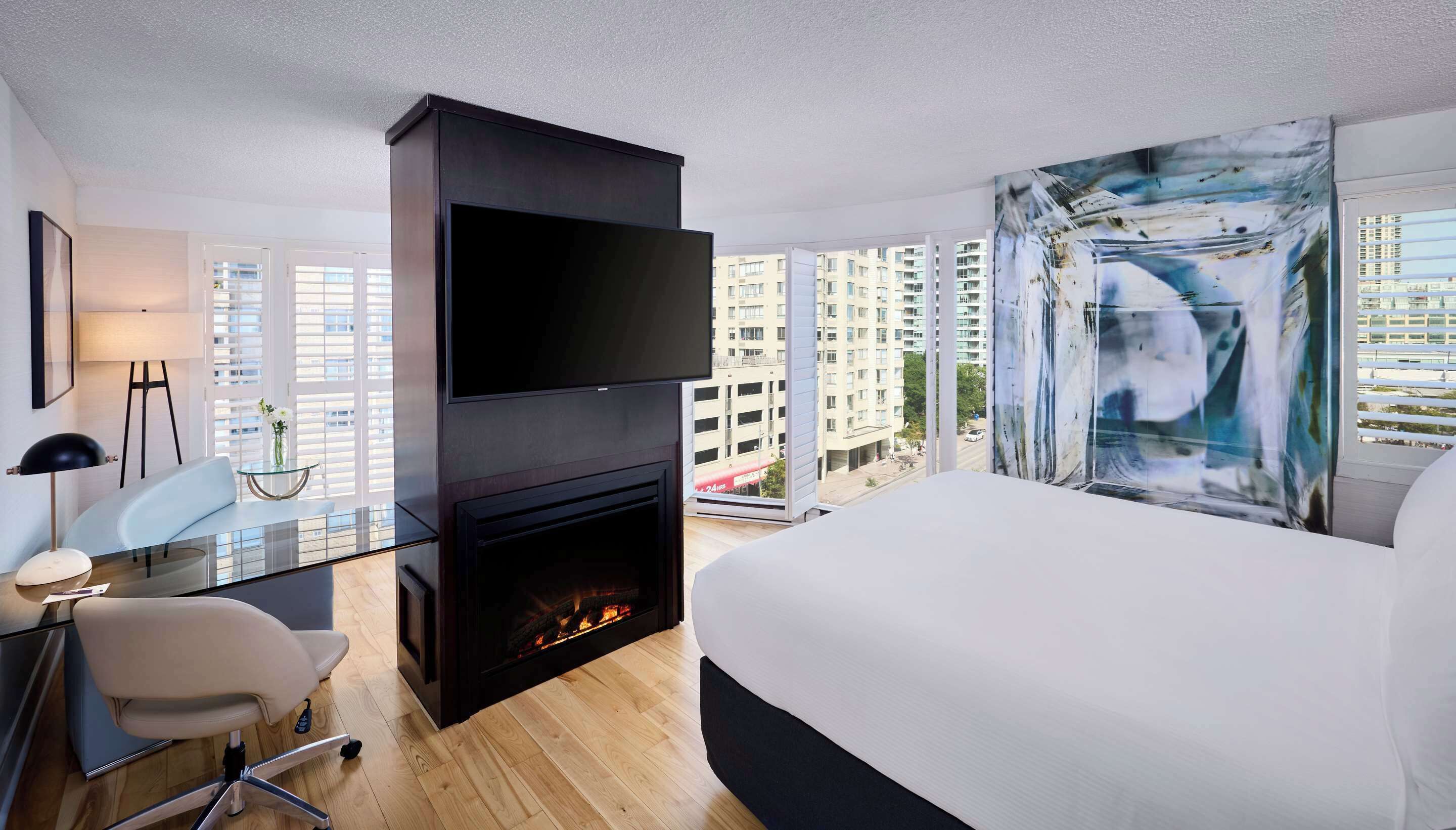 Quarto de hotel moderno com uma cama grande, televisão de tela plana, lareira, escrivaninha com cadeira e uma janela ampla com vista para a cidade.