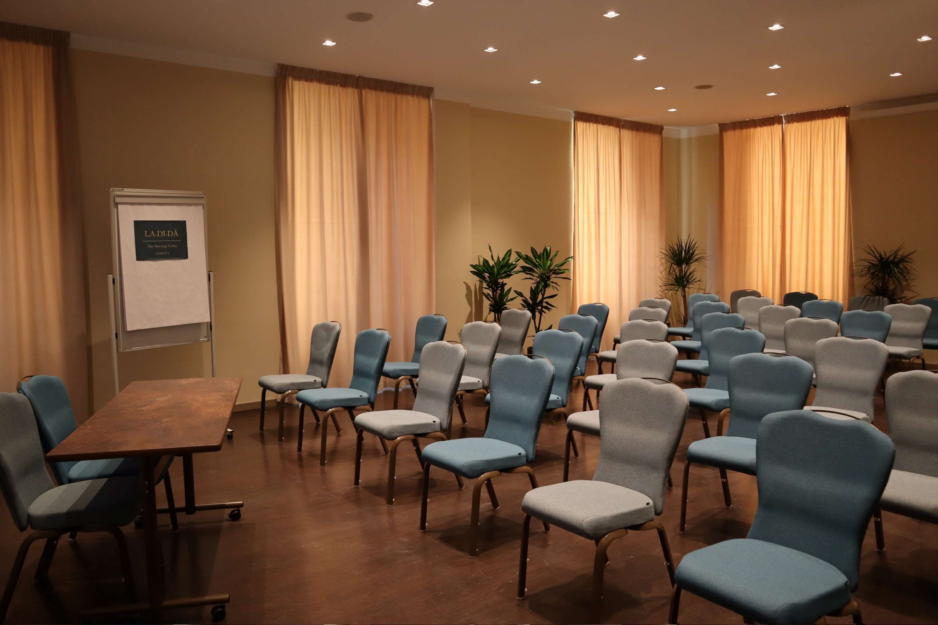 Uma sala de conferências com uma mesa de madeira e fileiras de cadeiras. Há duas janelas grandes com cortinas, e um aviso na parede.