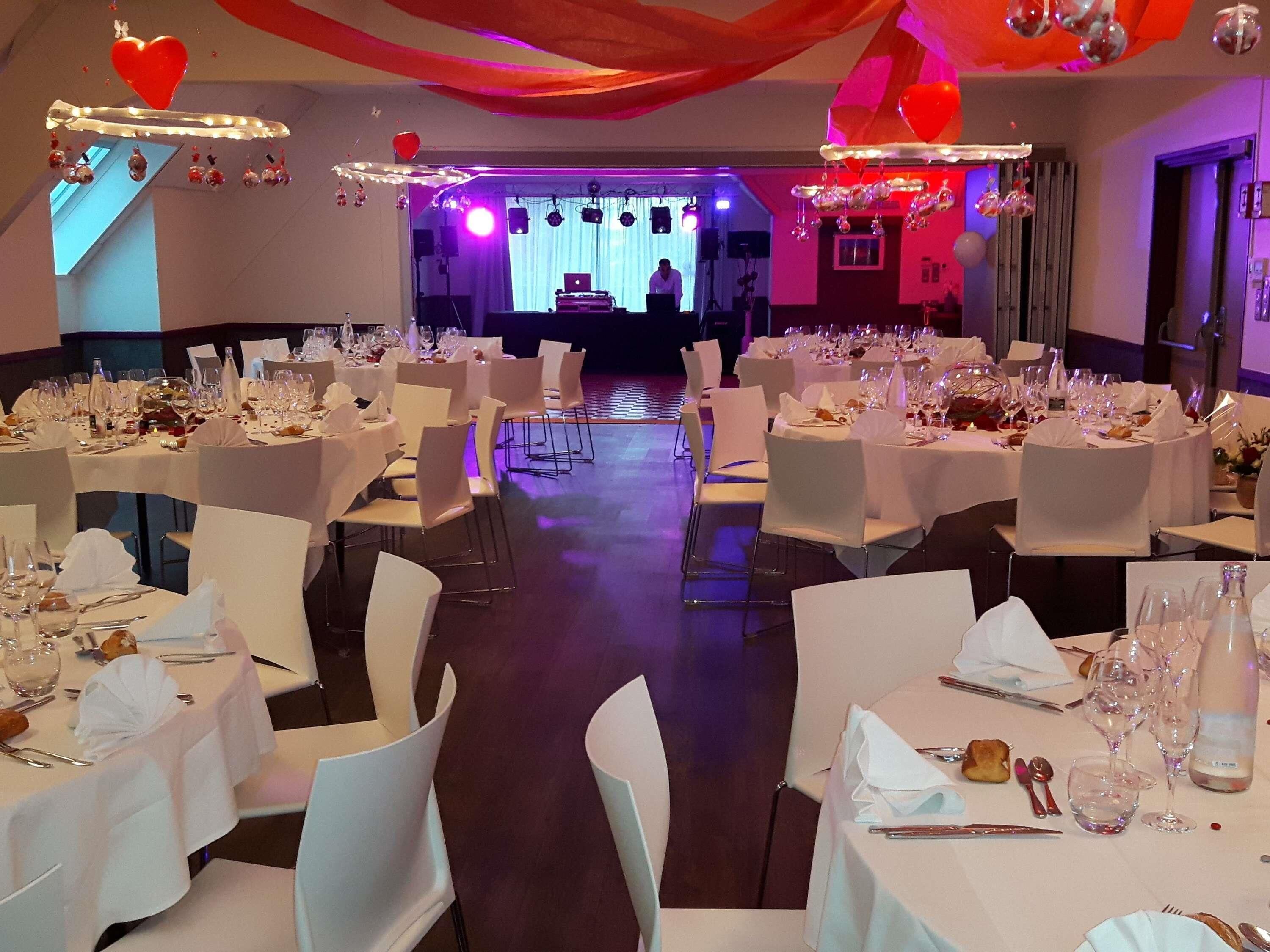 Um salão de banquetes com mesas montadas para um evento formal, com toalhas brancas, cadeiras e um palco com iluminação ao fundo.