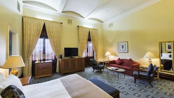 Quarto de hotel com uma cama grande, uma escrivaninha com televisão, um sofá vermelho, duas poltronas e uma mesa pequena.