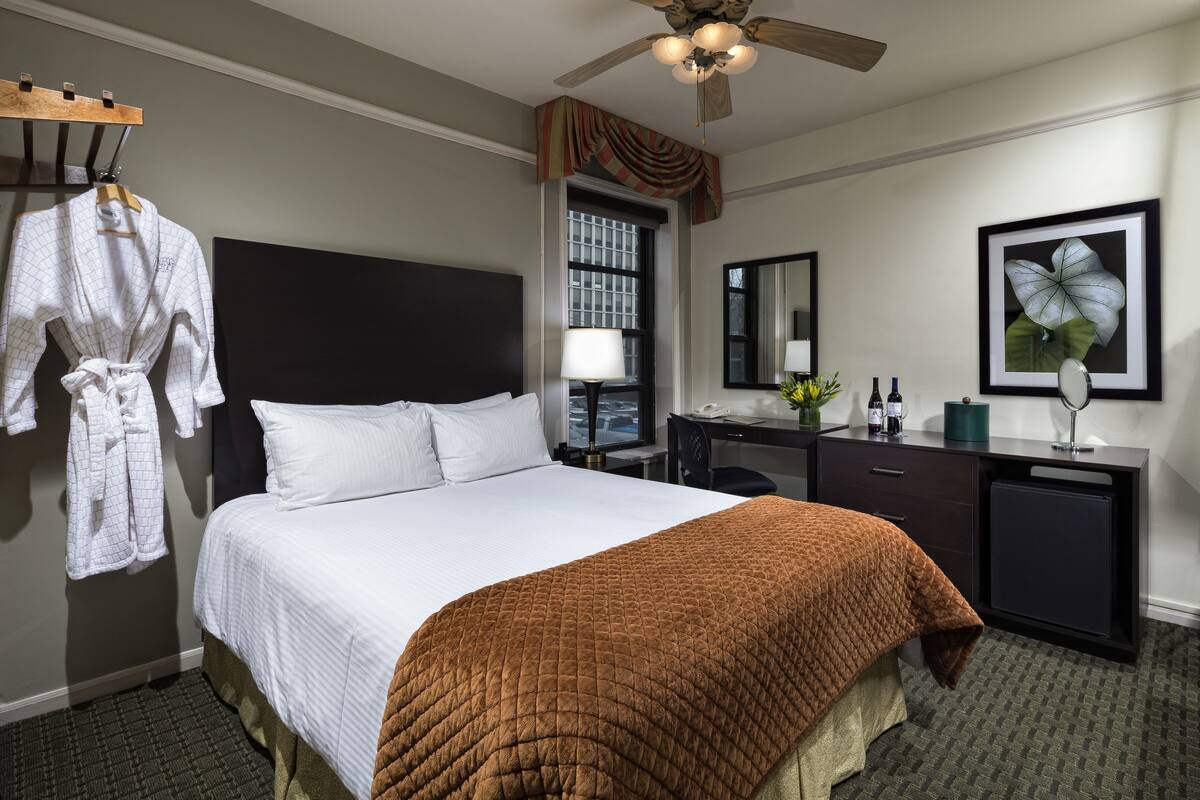 Quarto de hotel com cama, escrivaninha, cadeira, guarda-roupa, ventoinha no teto e janela com vista.