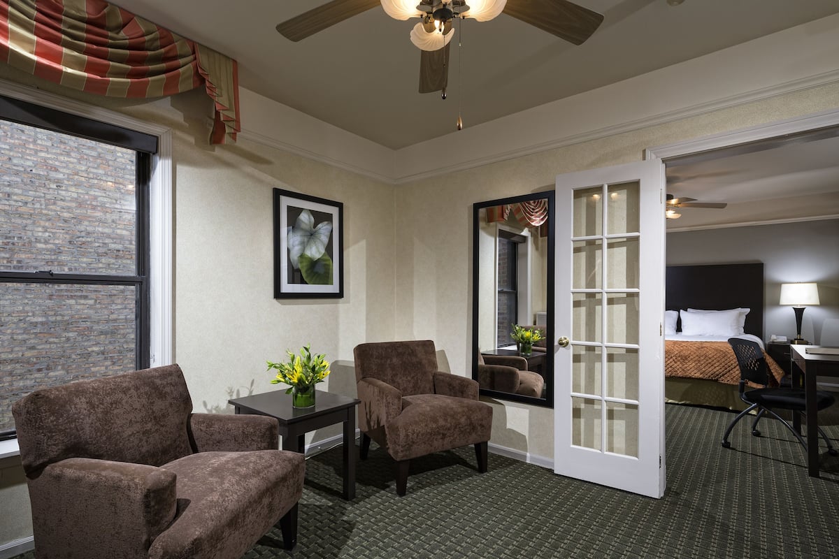 Quarto de hotel com parede de tijolos, dois poltronas, uma mesinha com uma planta, uma cama com edredom branco, uma escrivaninha com abajur e um ventilador de teto.