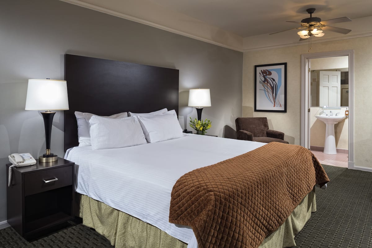 Quarto de hotel com uma cama grande, duas luminárias de cabeceira, uma mesa de cabeceira, uma cadeira, um quadro na parede e um banheiro visível através de uma porta aberta.