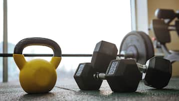 Um kettlebell amarelo e dois halteres pretos sobre o piso de uma academia, com uma janela ao fundo.